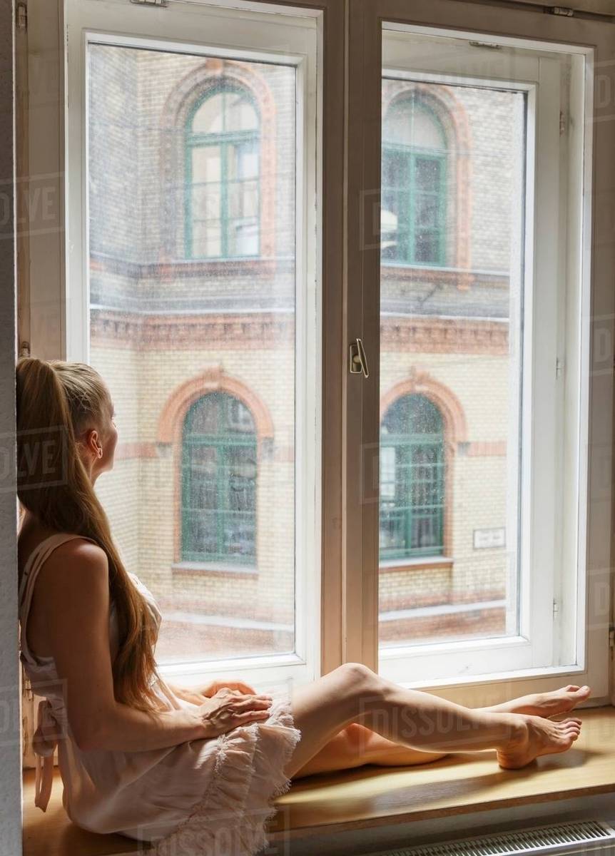 Mid adult woman sitting on window ledge, looking out of window, rear ...