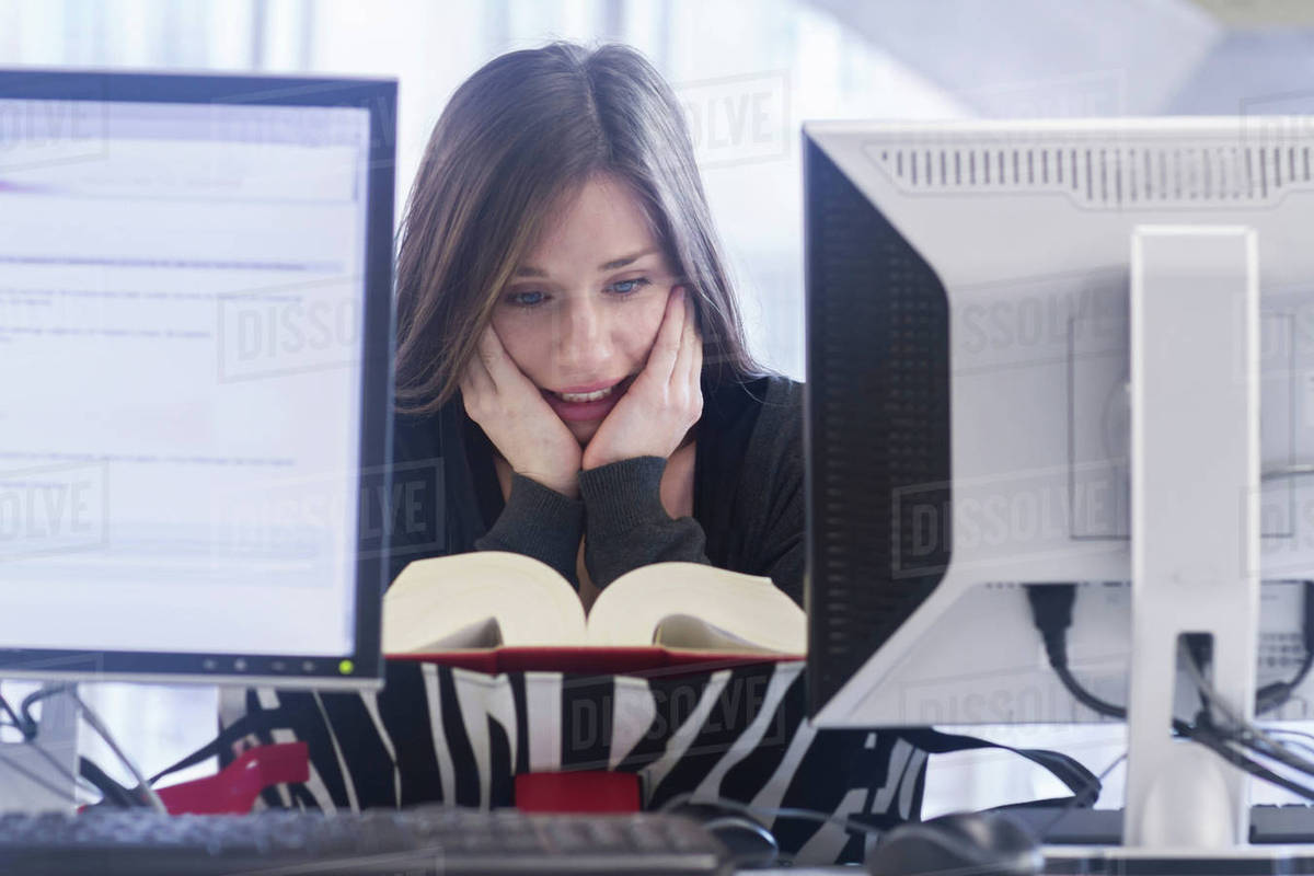 Woman sitting at computer chin in hand looking down reading book ...