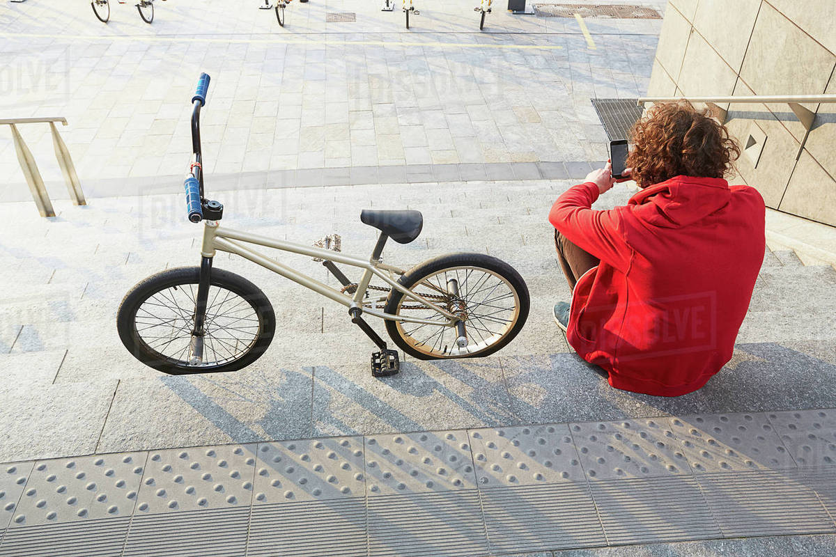 Rear view of man sitting on steps with BMX using smartphone - Royalty ...