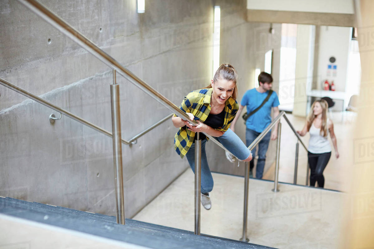 Young female student sliding down stairway handrail at higher education ...
