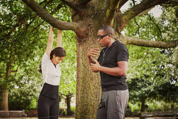 Personal trainer instructing woman on pull ups using park tree branch ...
