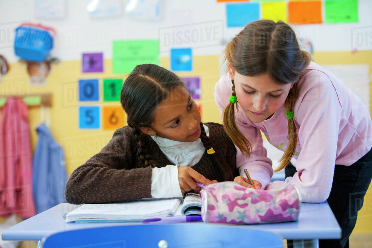 Primary schoolgirl helping friend with workbook in classroom - Royalty ...