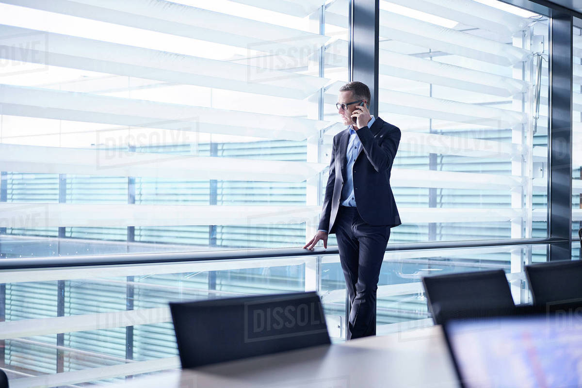 Businessman making smartphone call and looking through office window ...