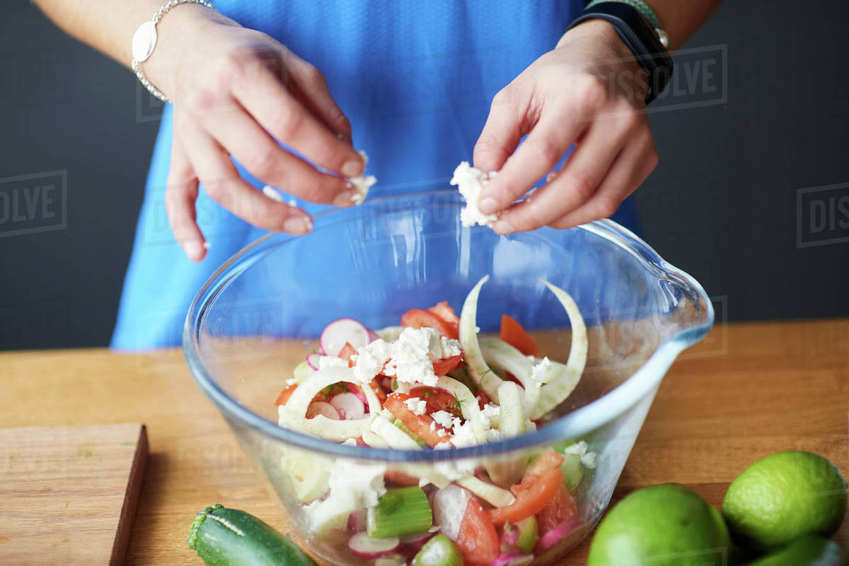Hands of young woman at kitchen table crumbling feta into salad bowl ...