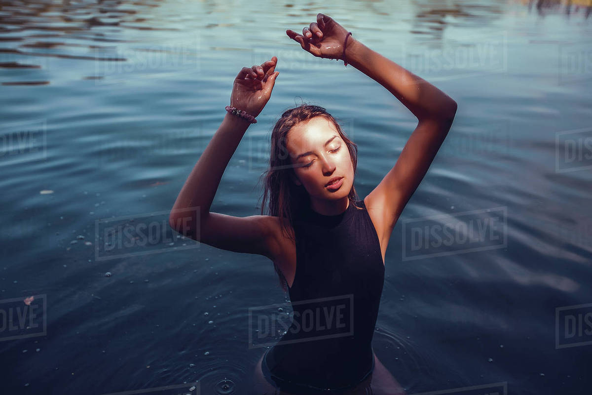 Portrait of woman wearing swimsuit standing in water, arms raised, eyes ...