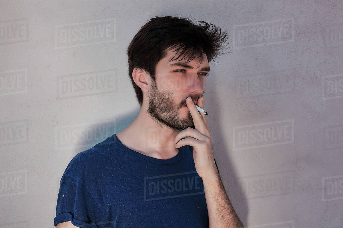 Young man with beard smoking cigarette in front of cement wall ...