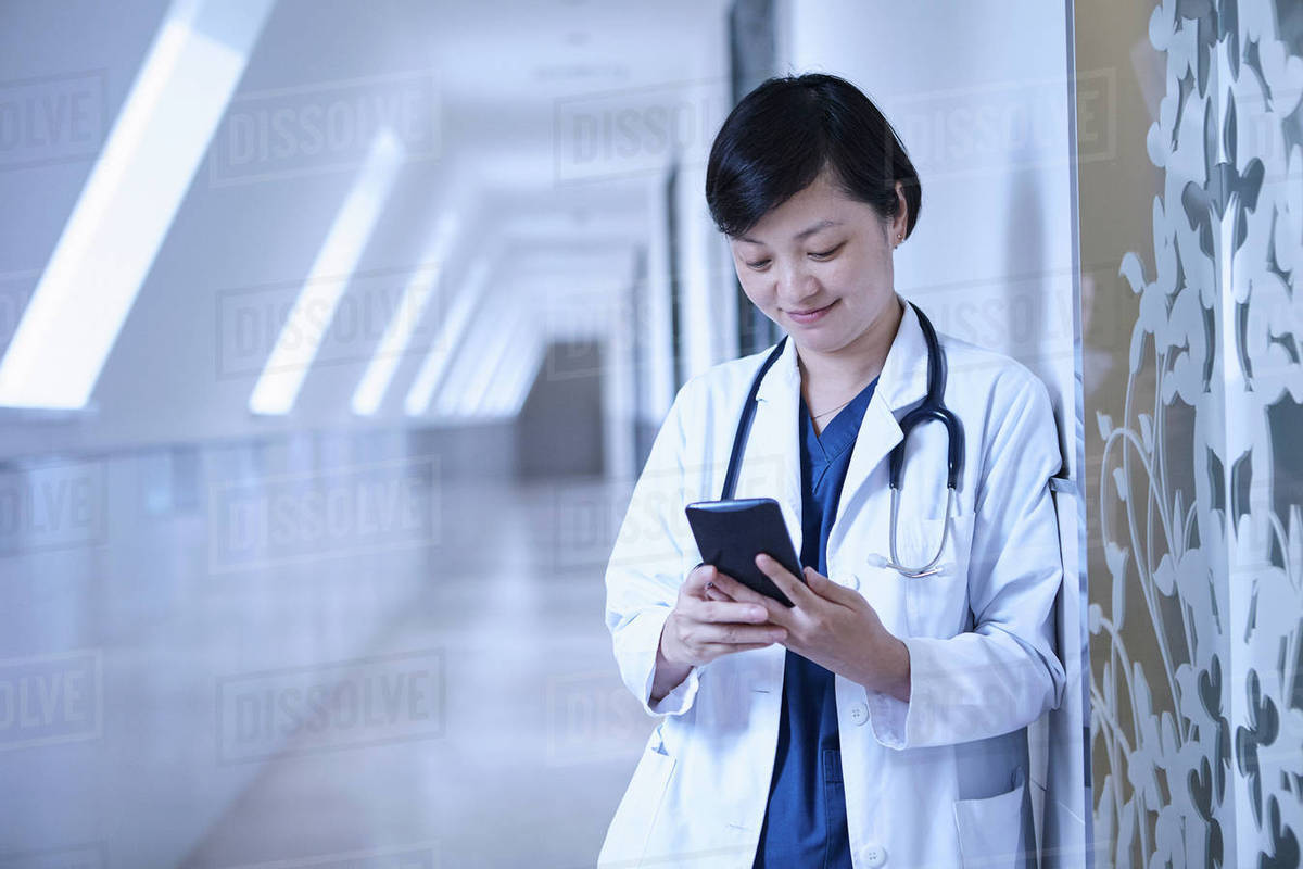 Doctor in hospital corridor leaning against wall using smartphone ...