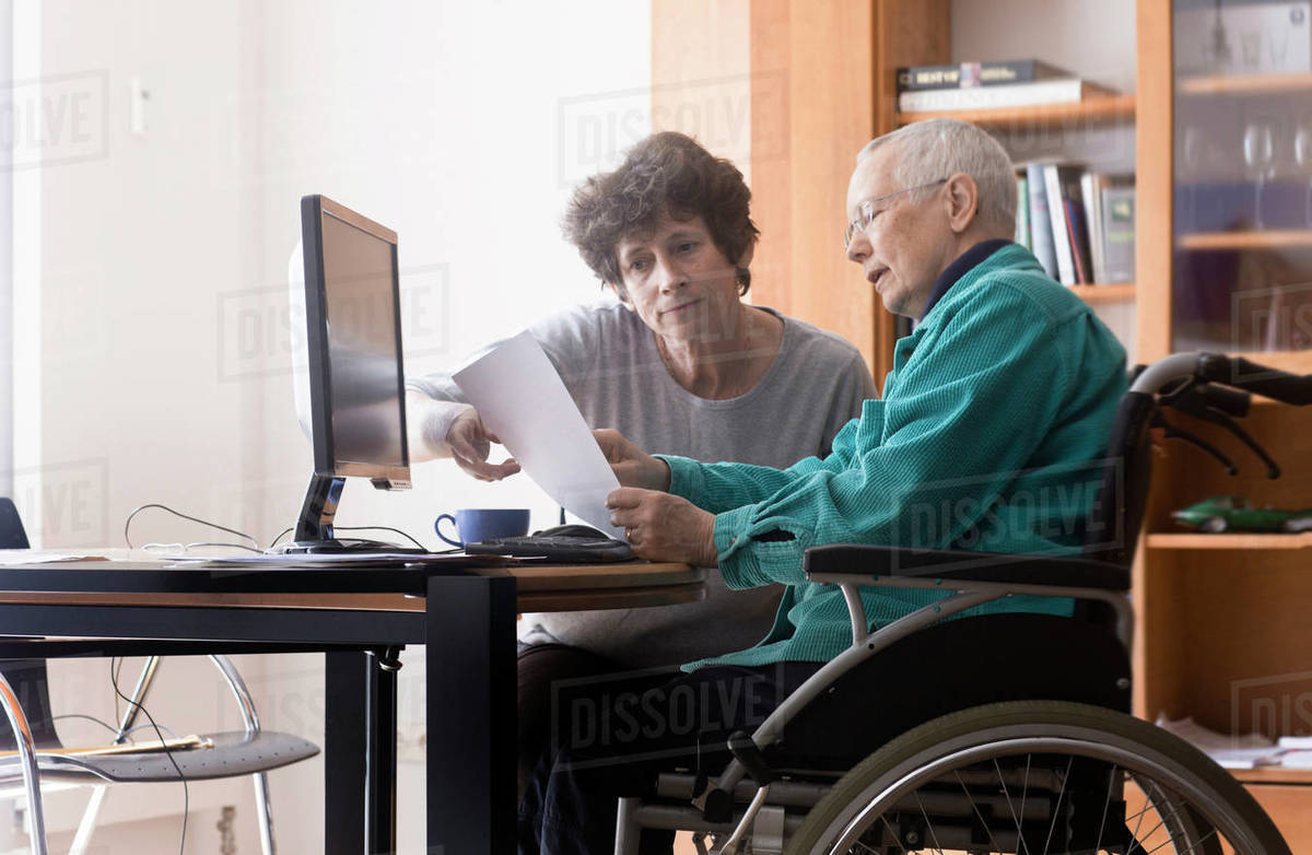 Senior woman sitting in a wheelchair at a computer, helper assisting ...