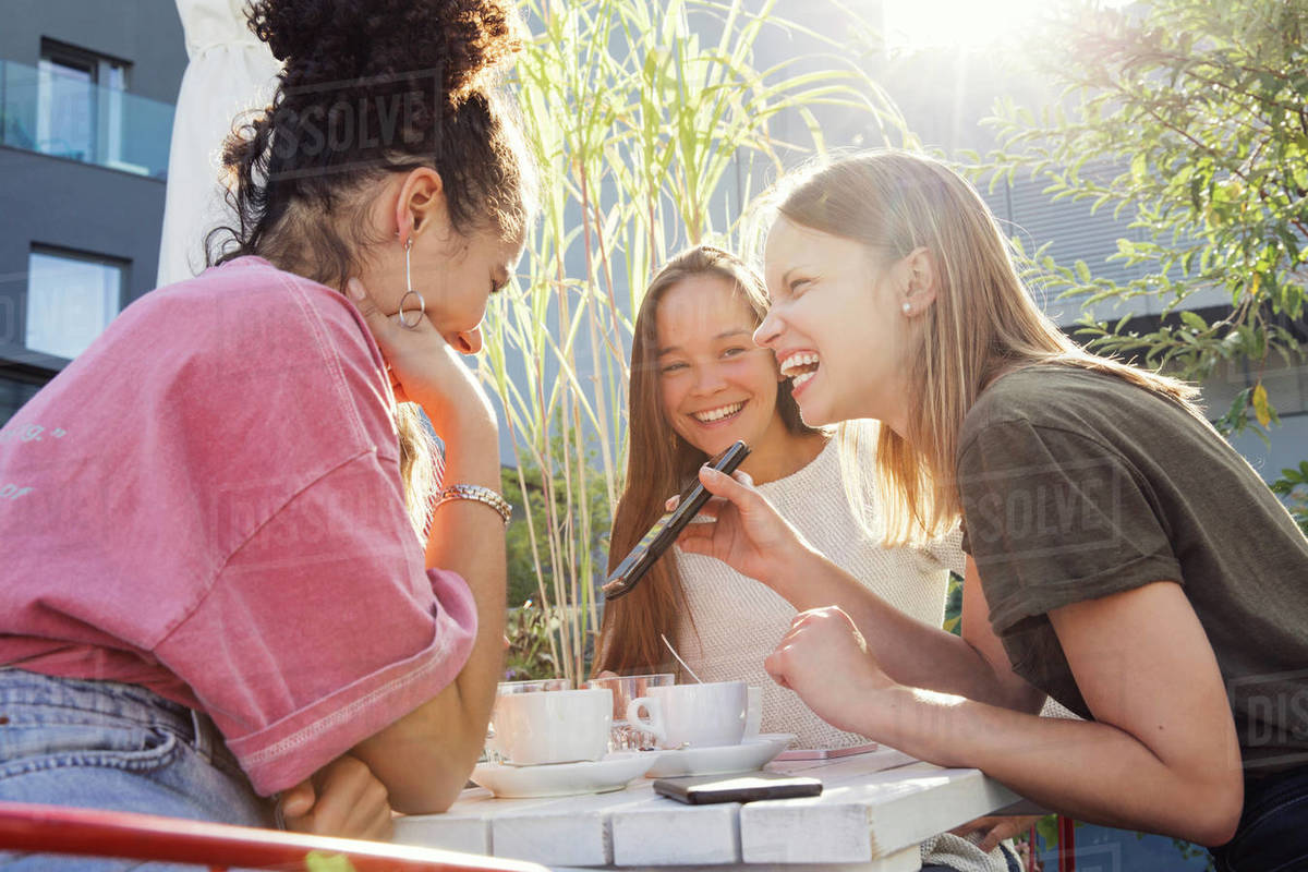 Three women sitting round a table looking at a mobile phone and smiling ...