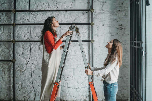 Two mid adult women on step ladders looking up in their new shop ...