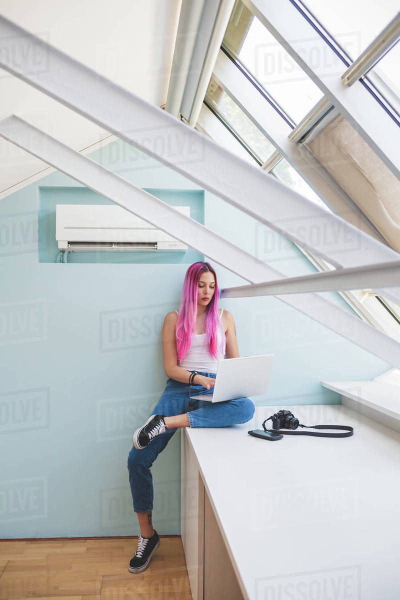 Young woman with long pink hair sitting on desk, typing on laptop ...