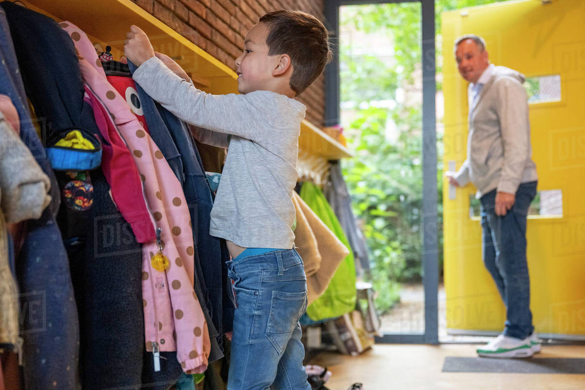 Young boy hanging up his coat at school with a teacher in the ...