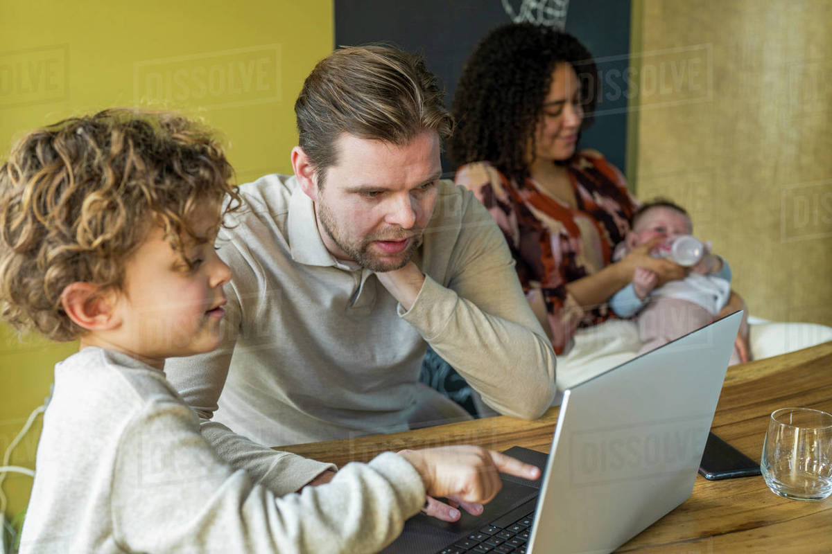 Father and child focused on a laptop while mother feeds a baby in the ...
