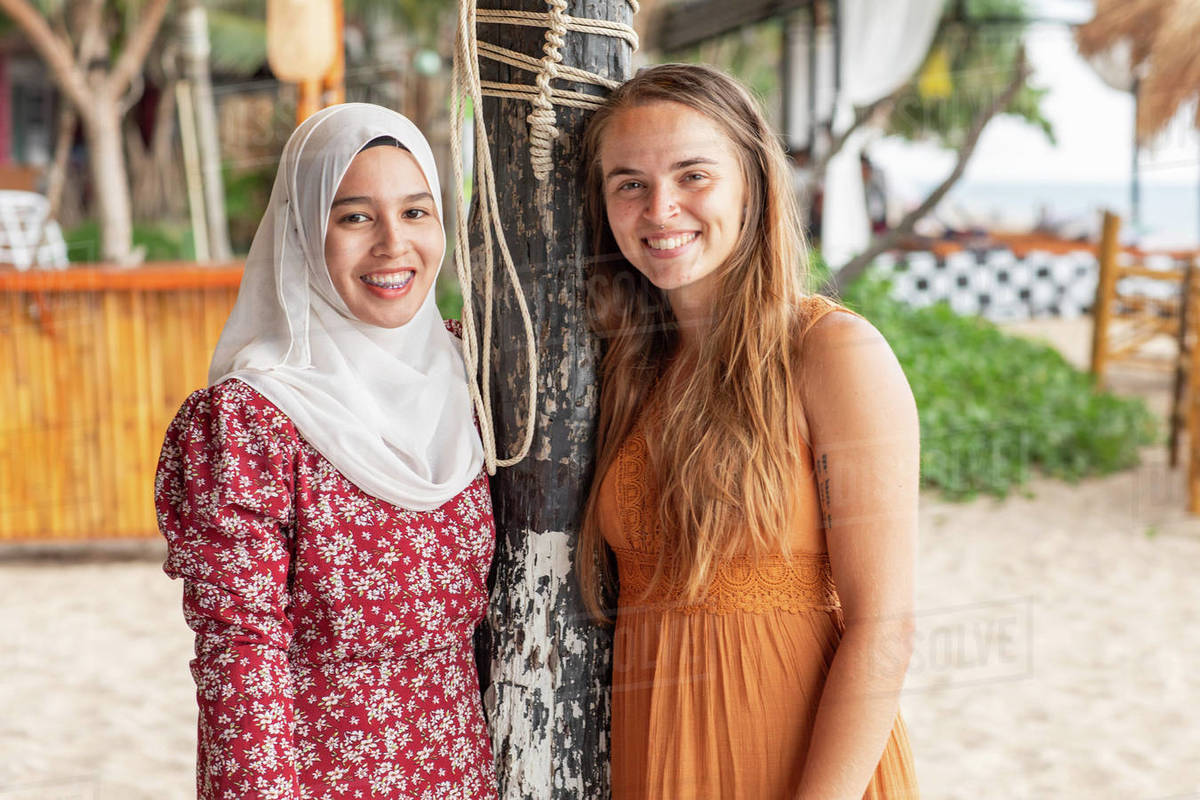 Portrait of two female best friends, one is wearing a hijab in a beach ...
