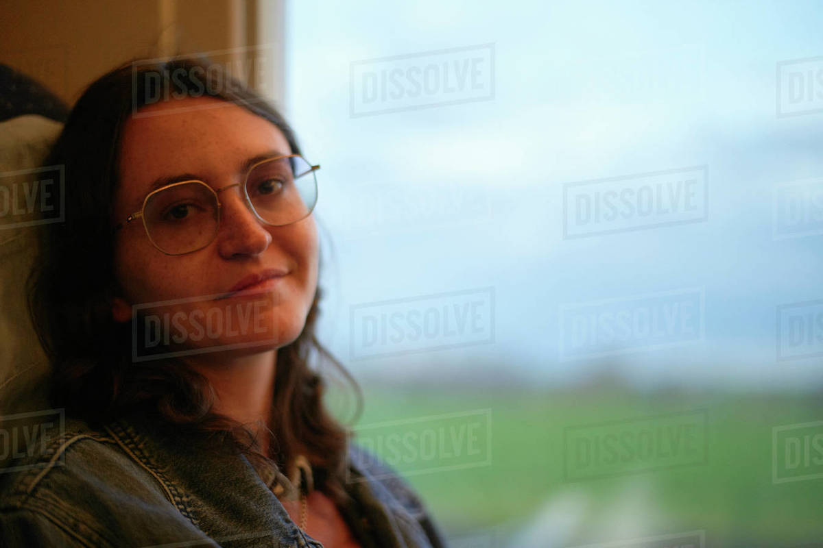 A woman with glasses is seated indoors by a window with a view of a ...