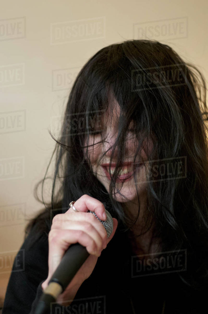 A person with long black hair is singing into a microphone with their ...