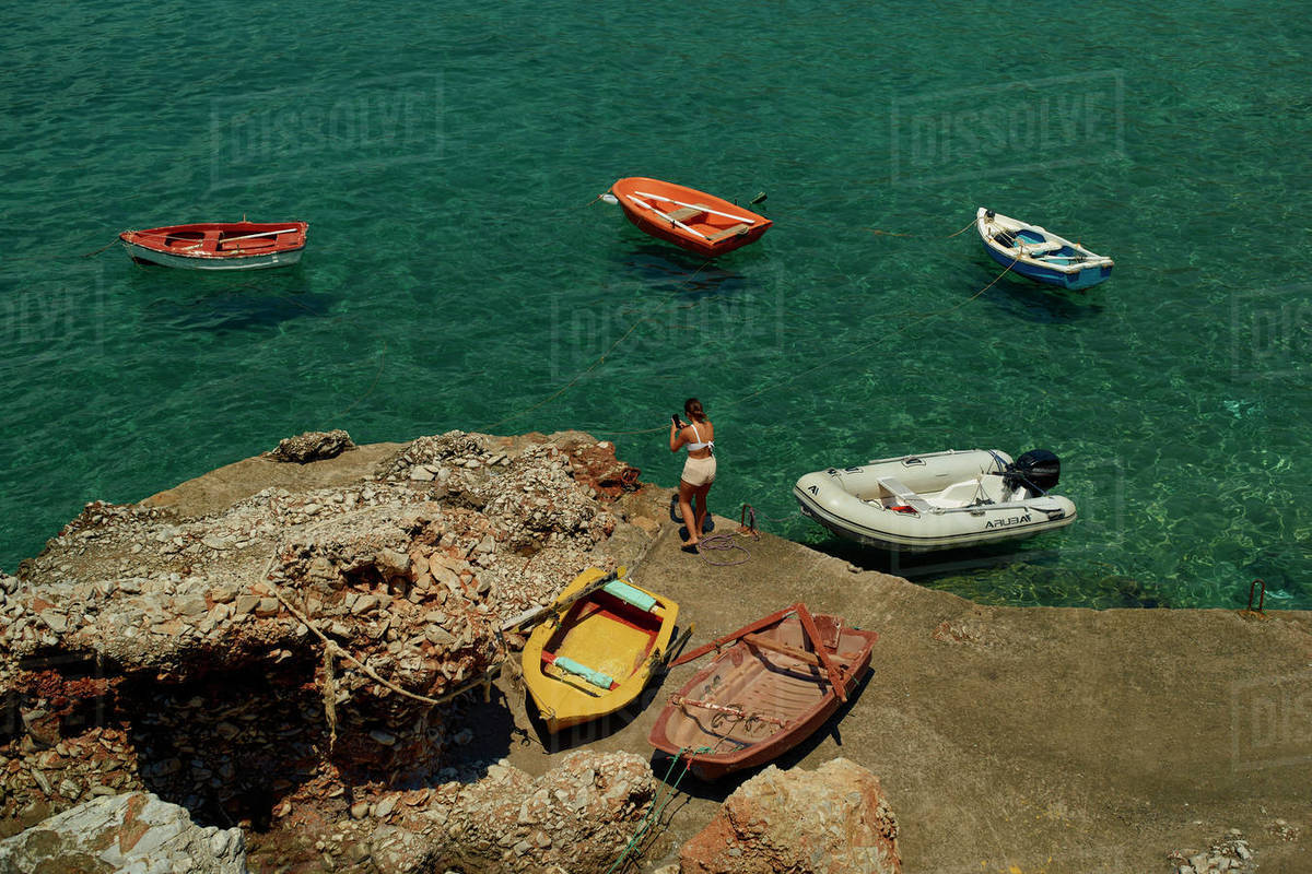 A person stands on a rocky shore beside colorful boats, overlooking the ...