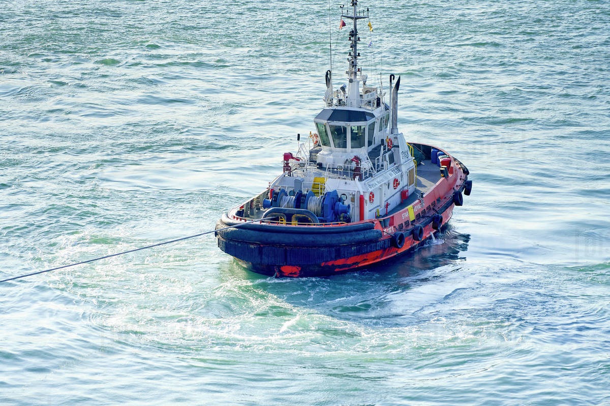 Red and blue tugboat towing a large unseen vessel on the open sea ...