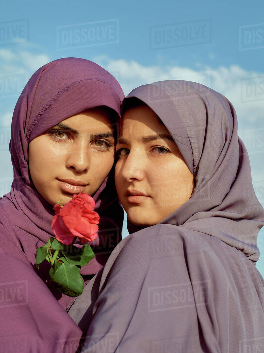 Two women wearing purple hijabs pose with a rose against a blue sky ...