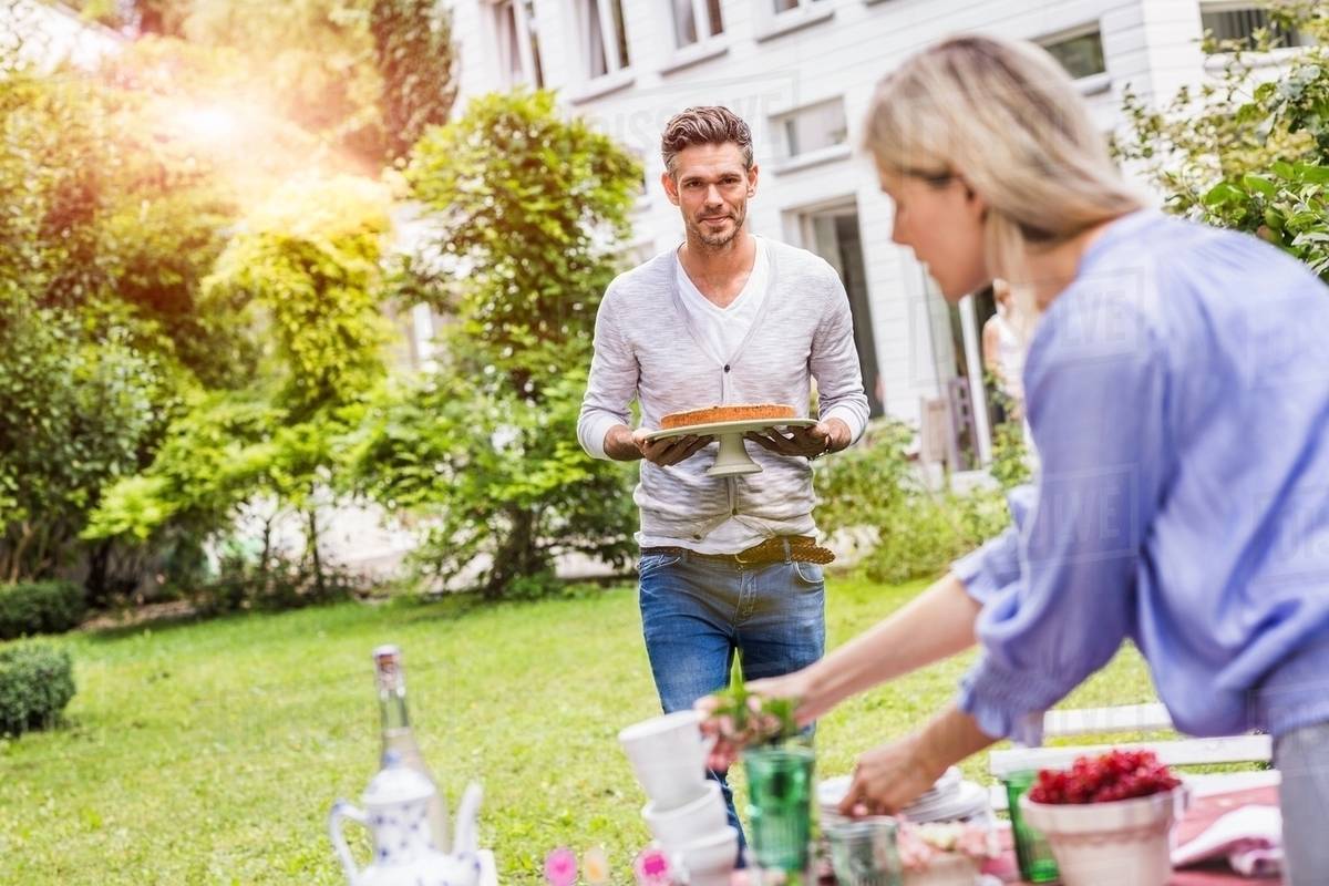 Mid adult man carrying cake on cake stand, walking towards garden table ...