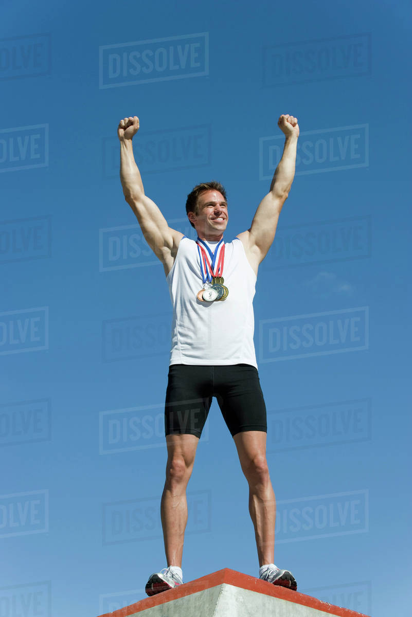 Male athlete standing on winner's podium with arms raised in victory ...