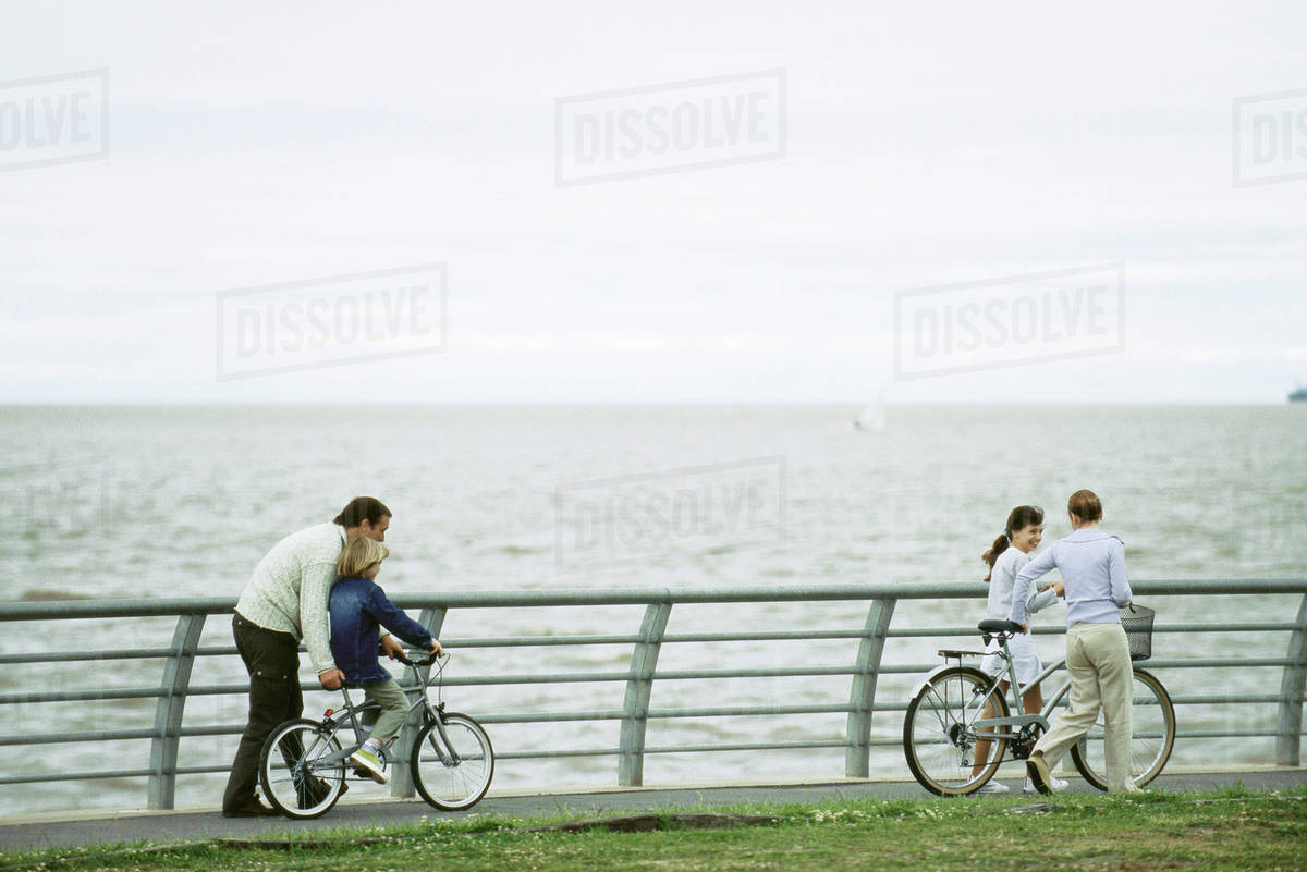 Family riding bicycles together at seaside park, father helping son ...
