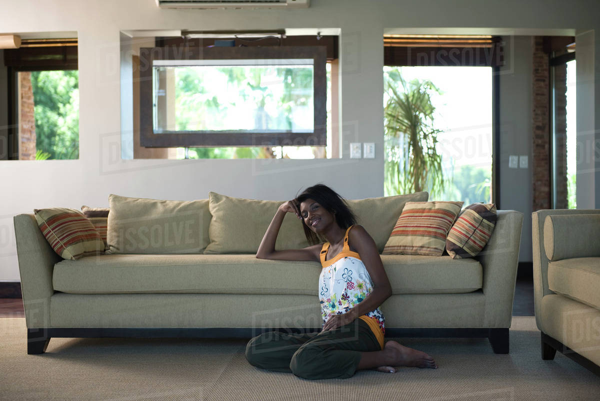 Young woman sitting on living room floor leaning against sofa, smiling ...