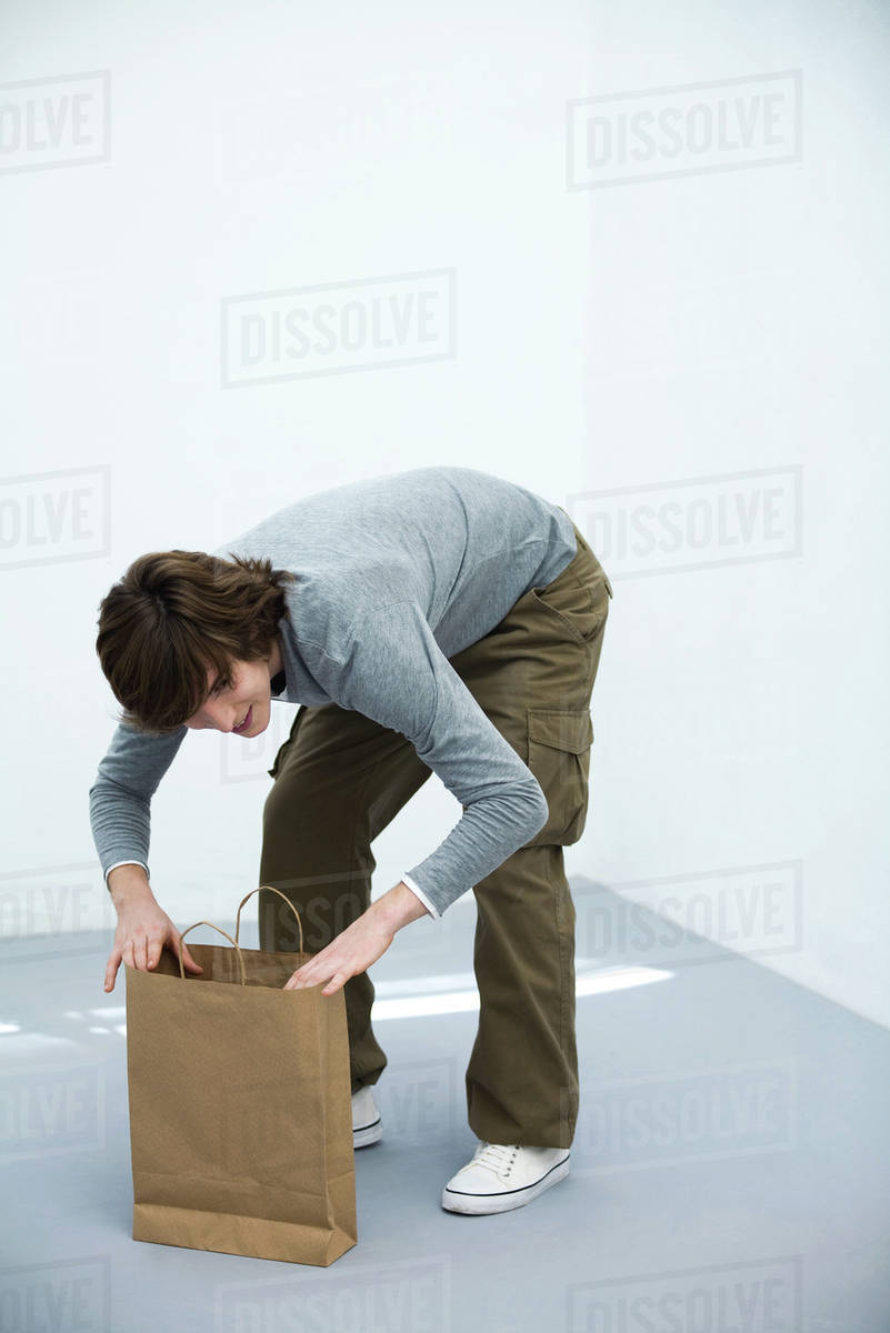 Young man bending over paper shopping bag, looking away, smiling ...