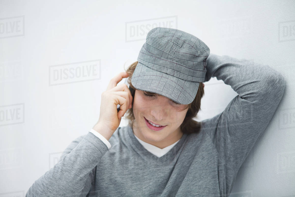 Young man in cap using cell phone, looking down, one hand behind head ...