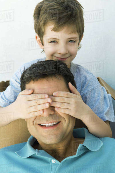 Boy with hands over his father's eyes, looking at camera, both smiling ...