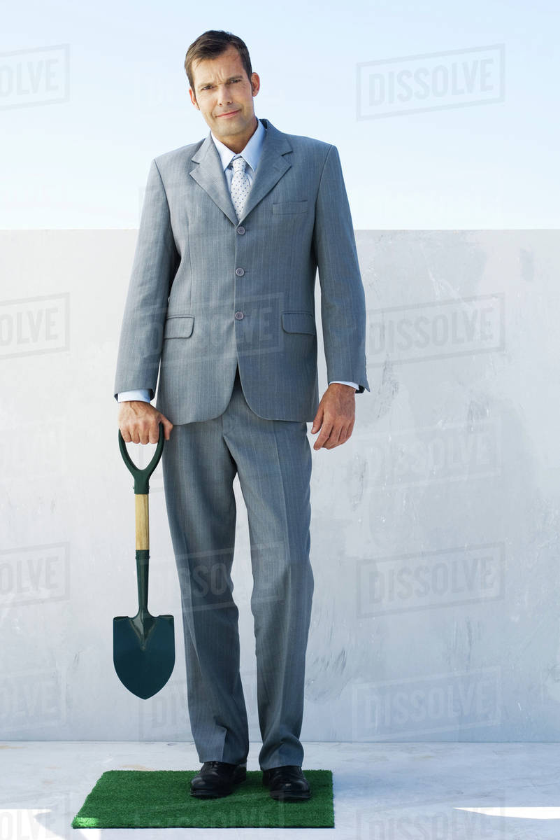 Man in suit, standing on small patch of artificial turf, holding shovel ...