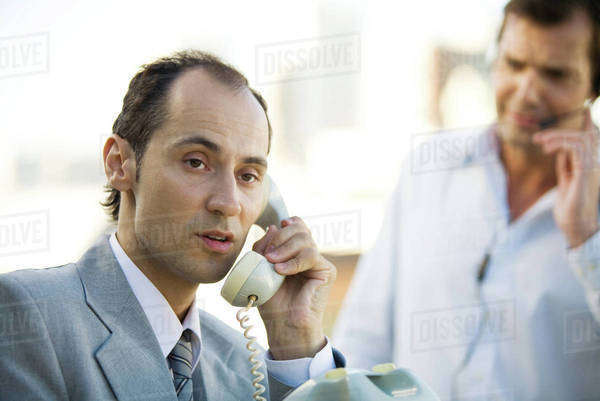 Businessman using landline phone, man in background using headset ...