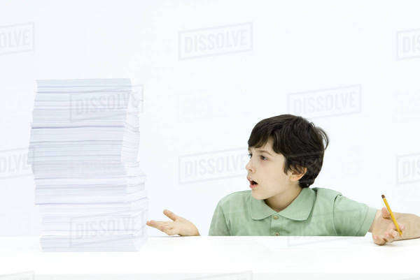 Boy looking at tall stack of paper, shrugging in disbelief, holding ...