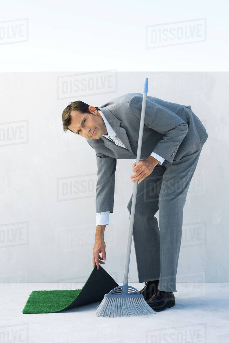 Businessman sweeping, lifting corner of rug, looking at camera ...
