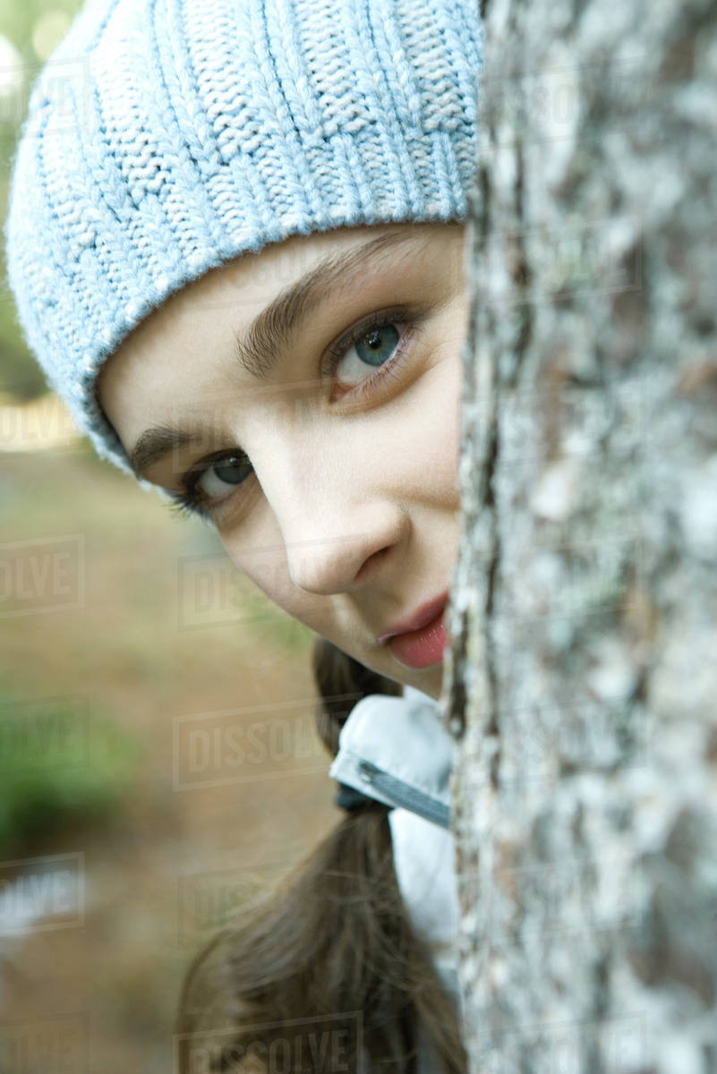 Teenage girl peeking around tree trunk, looking at camera, portrait ...
