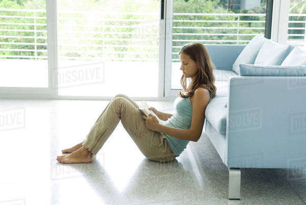 Teenage girl sitting on the ground, leaning against sofa, reading book ...