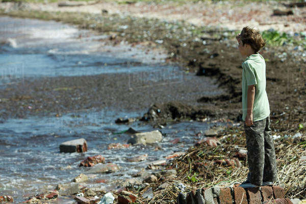 Boy standing on shore, looking out at polluted water - Royalty-free ...