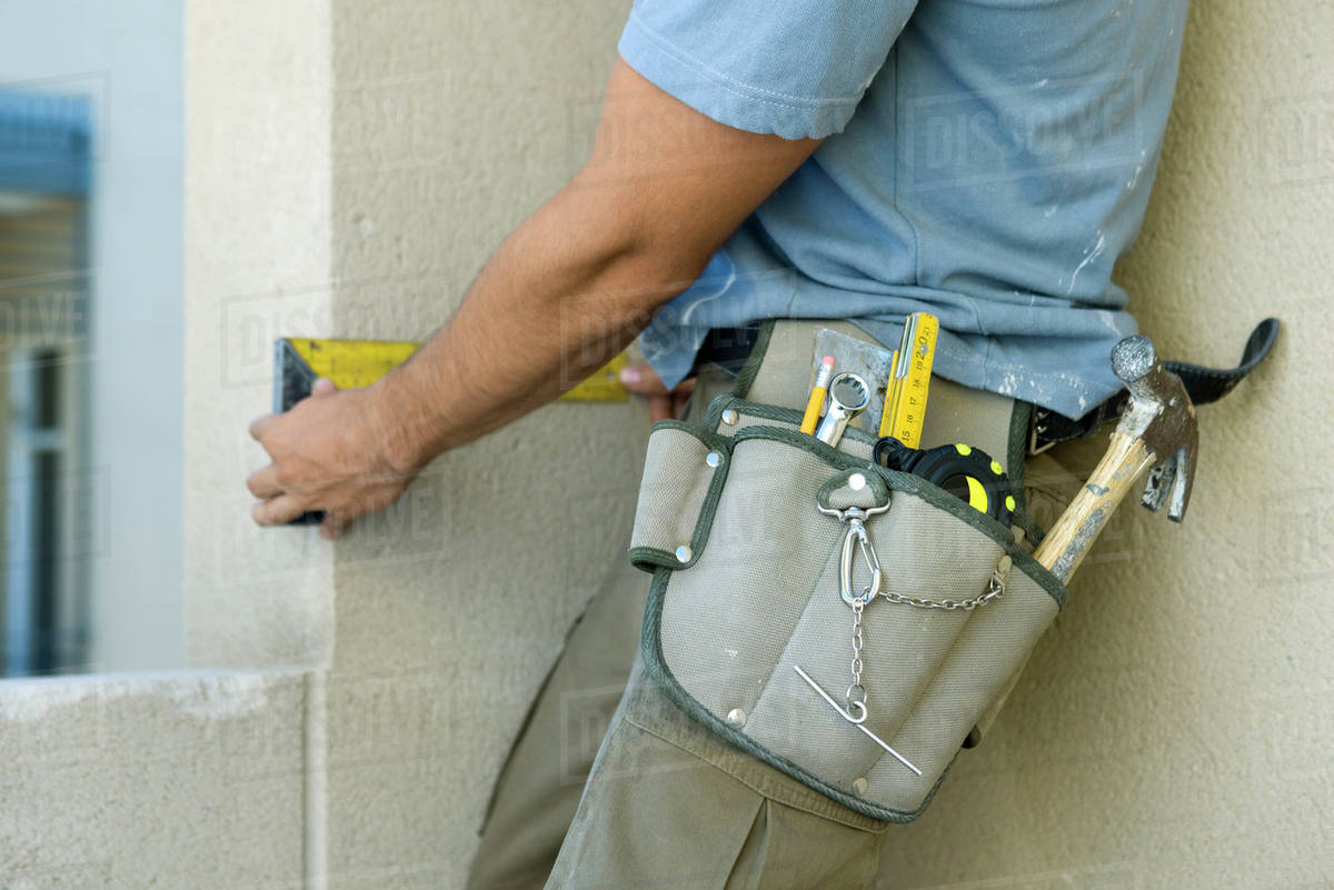 Construction worker wearing tool belt heavy with tools measuring wall ...