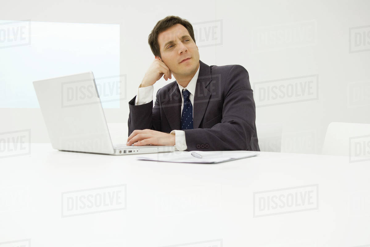 Businessman using laptop computer at desk, holding head, looking away ...