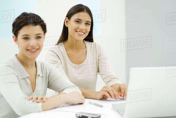 Two businesswomen sitting at desk together next to laptop computer, one ...