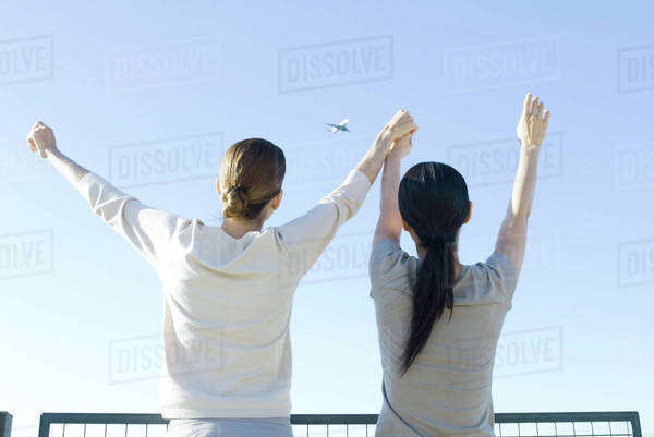 Two women looking at airplane in sky, holding hands, arms raised, rear ...