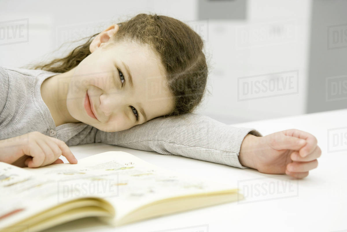 Girl sitting at table with book, resting head on arm, smiling at camera ...