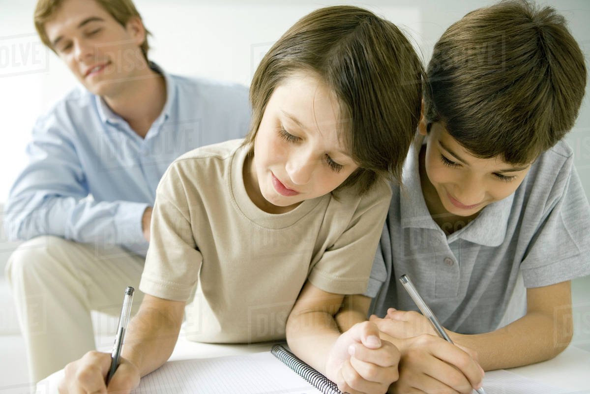 Two boys writing in notebook together, father watching in background ...