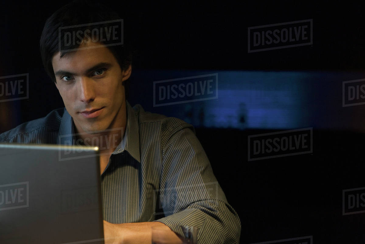 Man sitting in darkly lit room at table with laptop, looking at camera ...