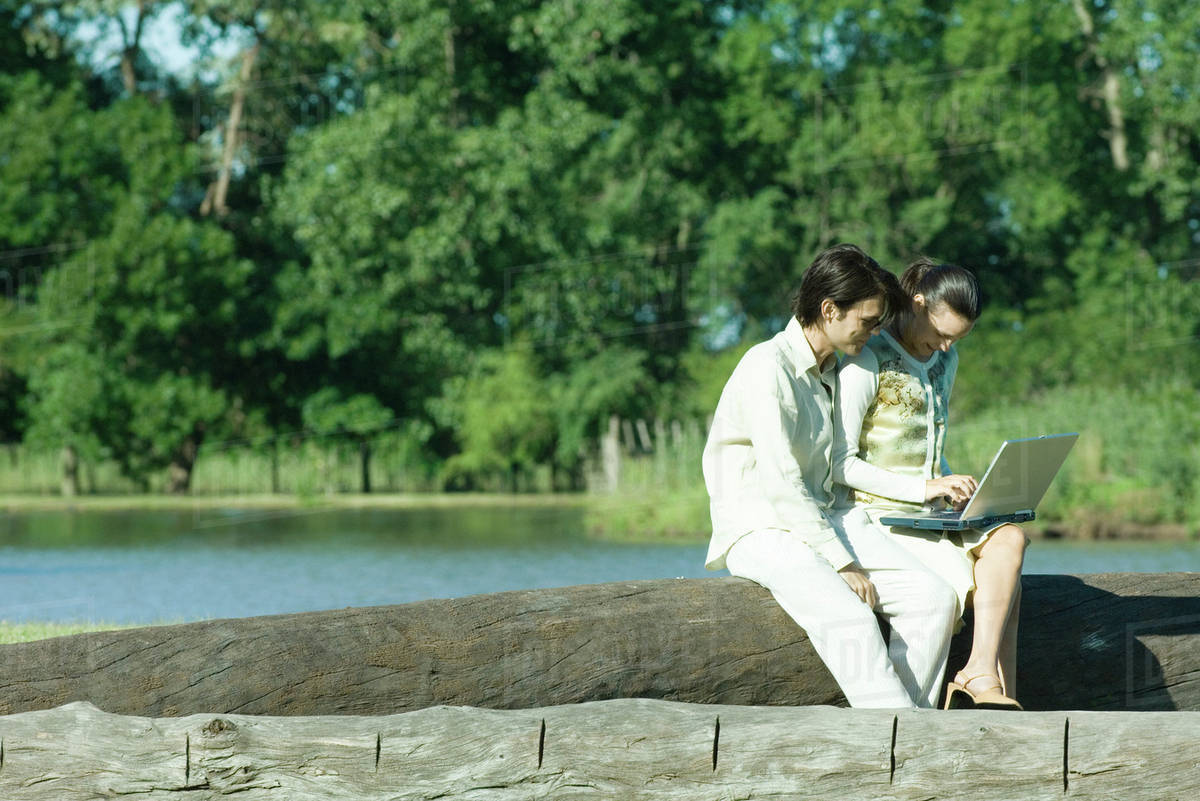 Couple sitting by water, using laptop - Stock Photo - Dissolve