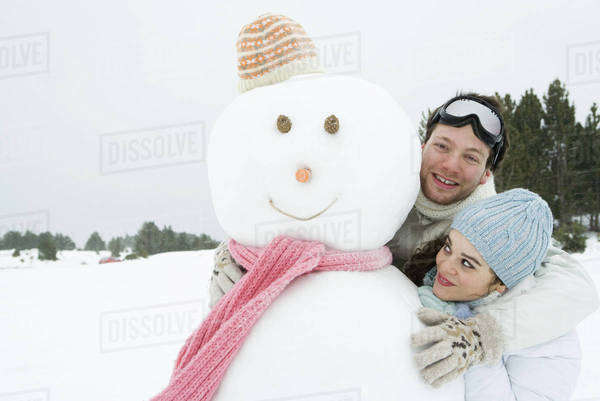 Two young friends standing with snowman, both smiling, one looking at ...