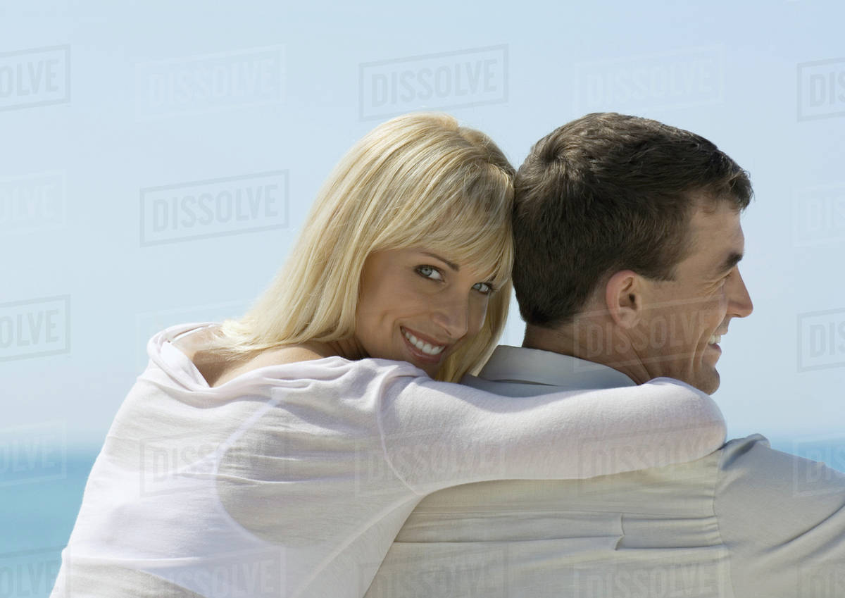 Woman leaning against man, looking over shoulder at camera, sea in ...