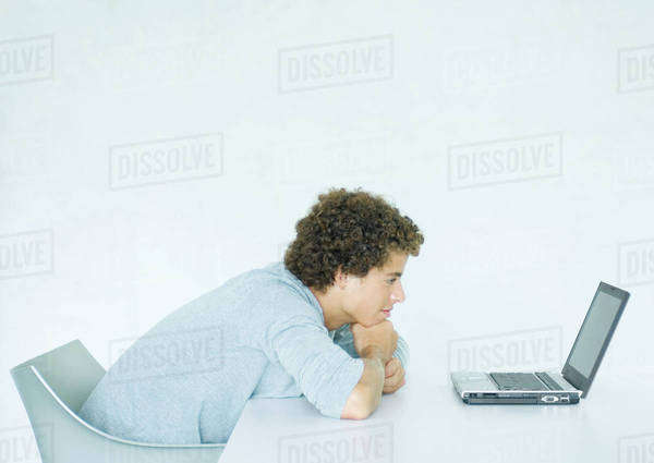 Young man sitting, leaning on desk, facing laptop computer - Royalty ...