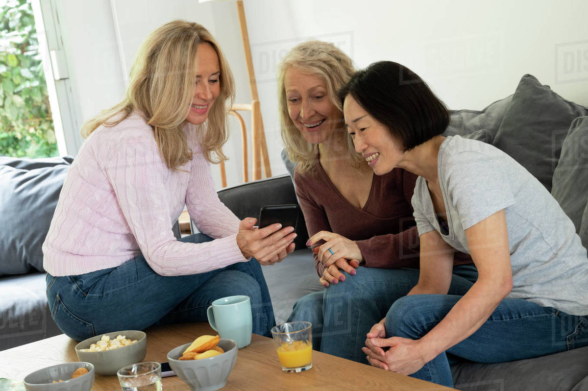 Portrait of three female middle-aged women having fun while looking at photos on phone in living ...