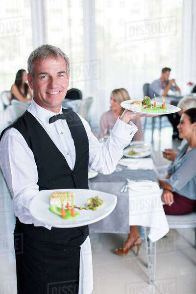 Portrait of smiling waiter holding fancy dishes, people sitting at ...